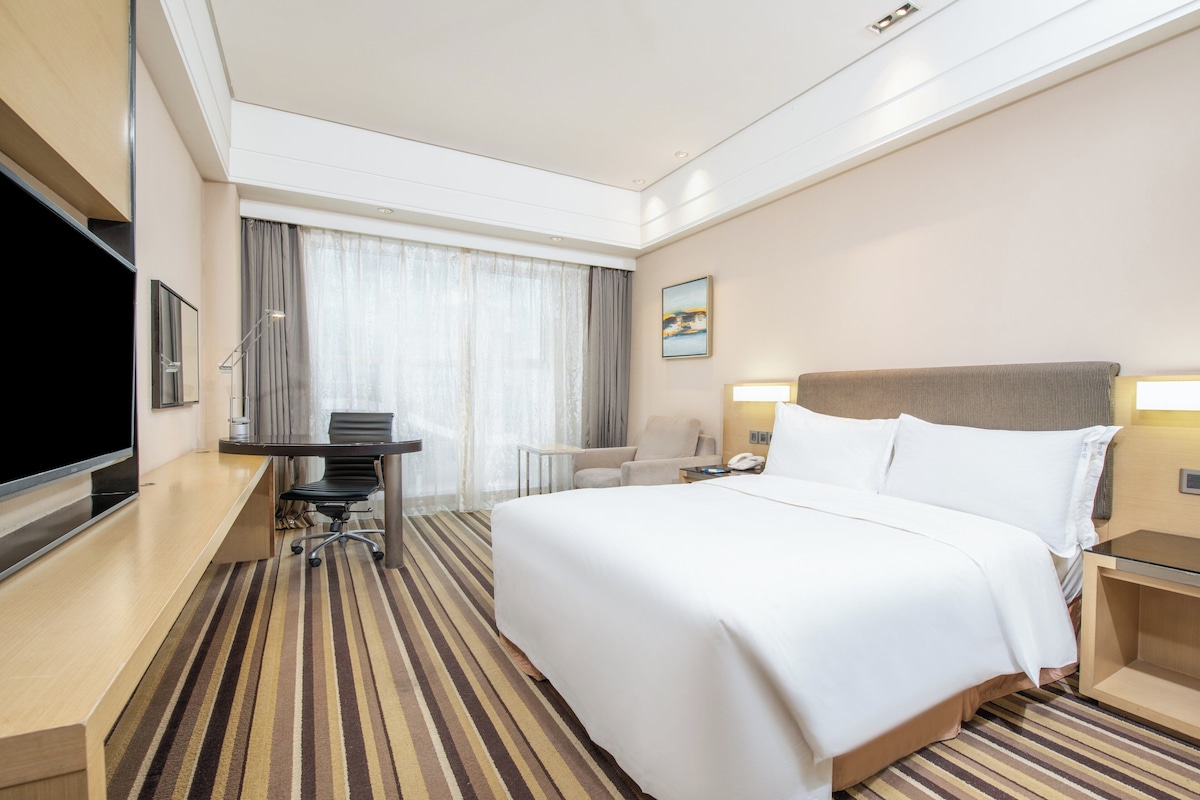 Quarto de hotel com cama grande, escrivaninha com cadeira, televisão e um quadro na parede.