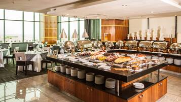 Buffet montado com diversas estações de serviço, grande variedade de comidas e vista para a cidade através de amplas janelas.