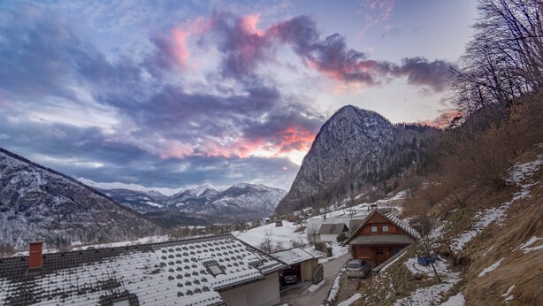 Uma aldeia montanhosa com telhados e edifícios cobertos de neve, uma montanha imponente ao fundo, e um céu claro com nuvens.