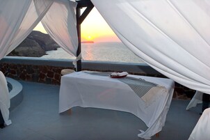 Uma cama com dossel branco com vista para um pôr do sol à beira-mar.