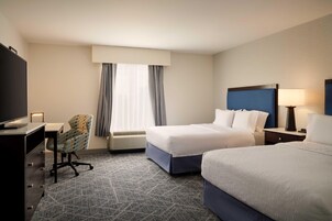 Quarto de hotel com duas camas, uma mesa de trabalho com cadeira, televisão e janela com cortinas.