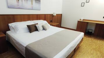 Uma cama bem arrumada com roupa de cama bege e branca, uma cabeceira de madeira, uma mesa de cabeceira com abajur, uma escrivaninha pequena e uma lixeira.