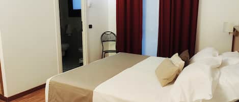 Quarto de hotel com cama, uma cadeira, um guarda-roupa e um banheiro visível através de uma porta aberta.