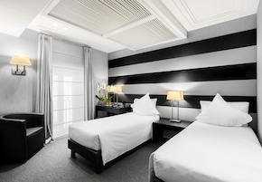Quarto de hotel com duas camas, uma escrivaninha, uma cadeira e uma janela grande com cortinas.
