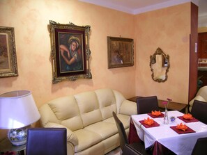 Uma área de refeições com um sofá bege, uma mesa posta com guardanapos vermelhos e um vaso de flores, e duas pinturas emolduradas na parede.