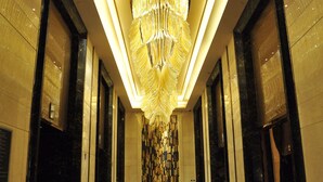 Um corredor longo e bem iluminado, com um lustre central, paredes de mármore e um tapete estampado.