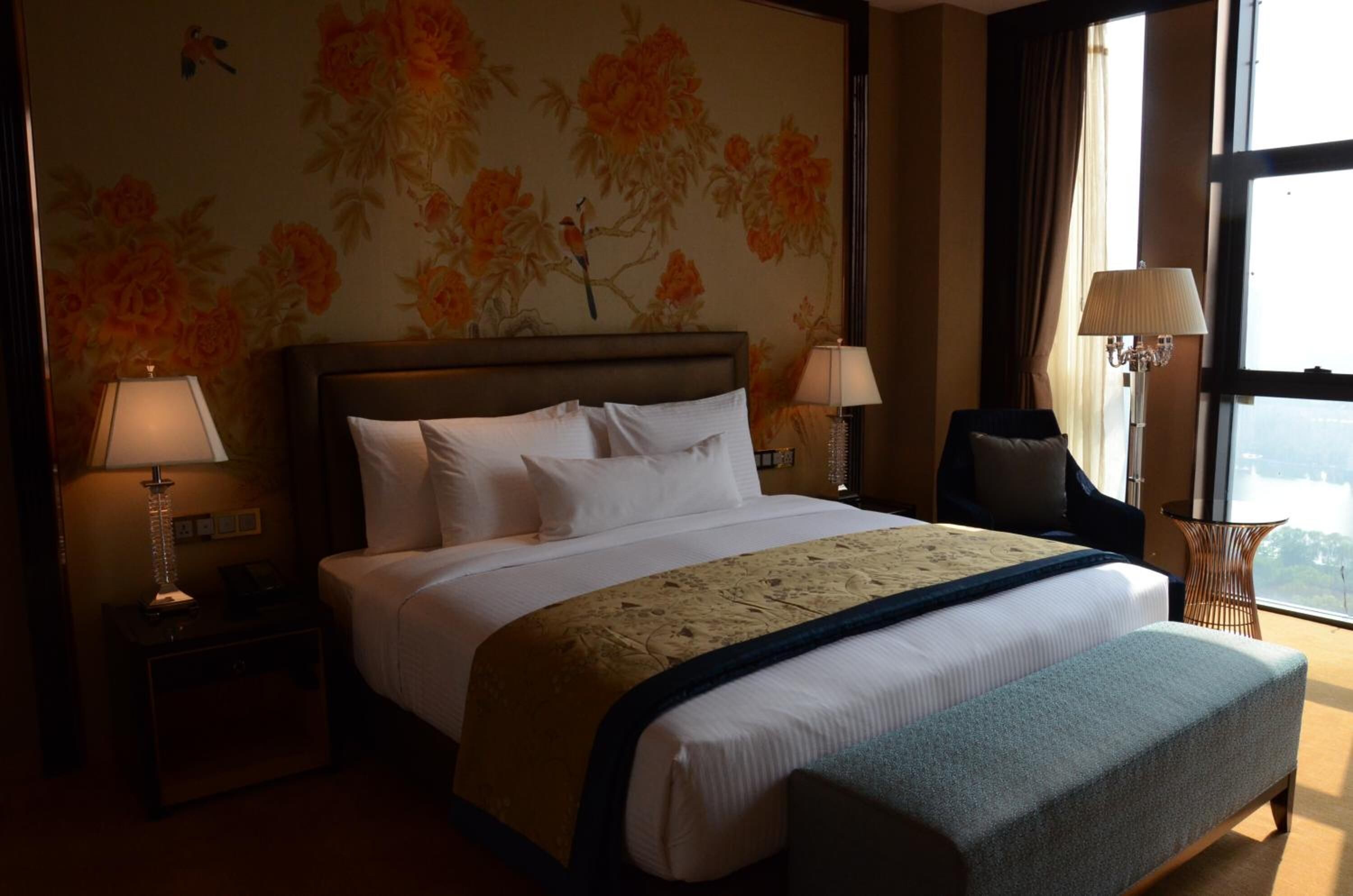 Quarto de hotel com uma cama grande, duas mesinhas de cabeceira com luminárias, um banco e vista para o exterior através de uma janela ampla.