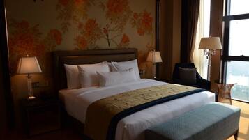 Quarto de hotel com uma cama grande, duas mesinhas de cabeceira com luminárias, um banco e vista para o exterior através de uma janela ampla.
