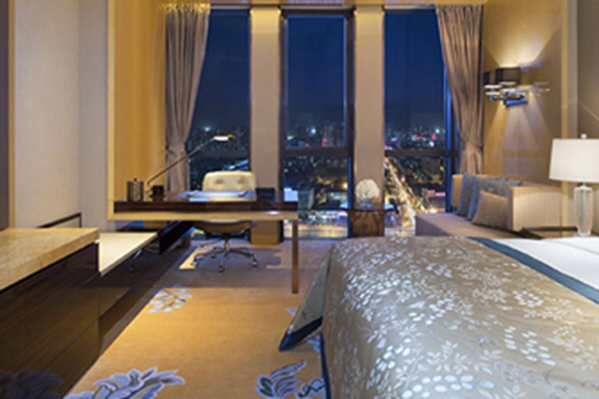 Quarto de hotel com cama, escrivaninha, cadeira, luminária e vista da cidade à noite.