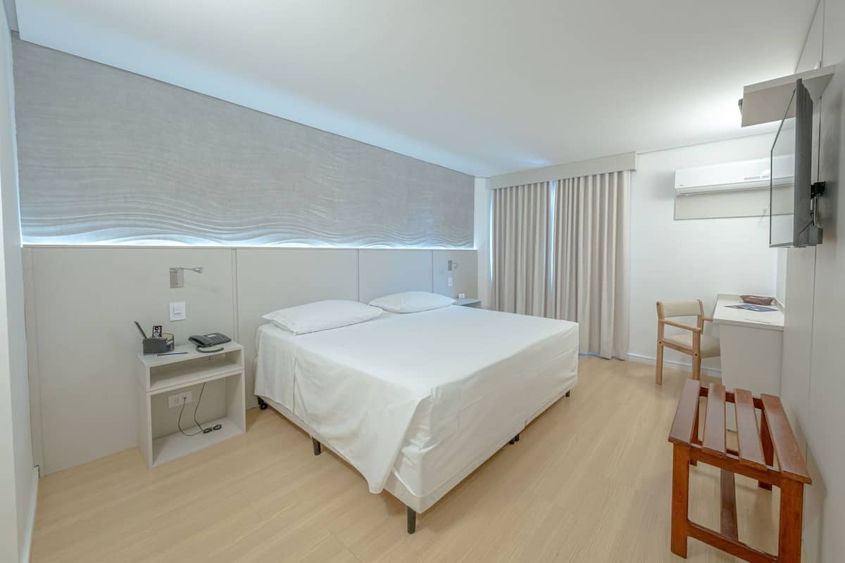 Quarto de hotel com cama, escrivaninha, cadeira e prateleira pequena com telefone e rádio.