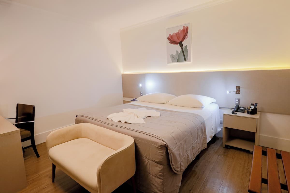 Quarto de hotel com uma cama, uma cadeira, mesa de cabeceira com telefone e abajur, e um quadro na parede.
