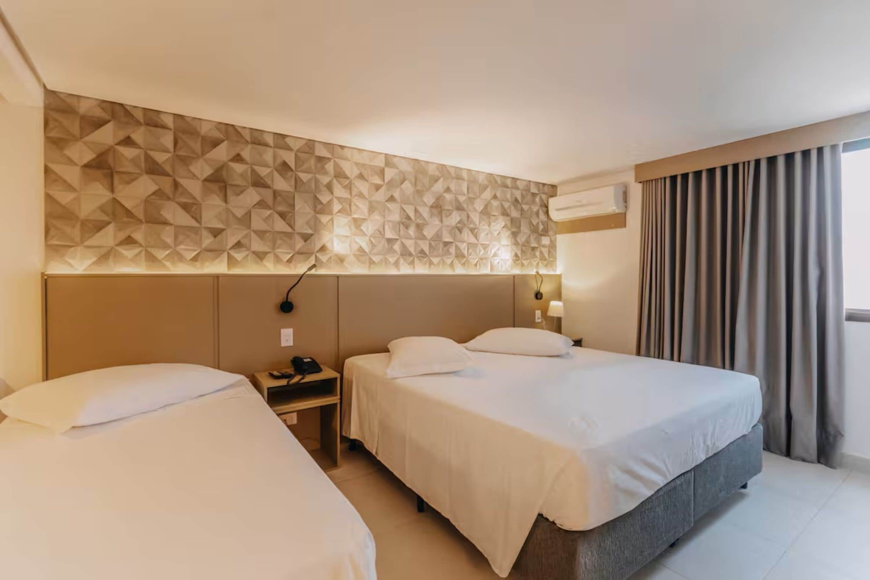 Quarto de hotel com duas camas, cabeceira estampada com padrão geométrico, luminárias laterais, mesa de cabeceira e janela com cortinas.