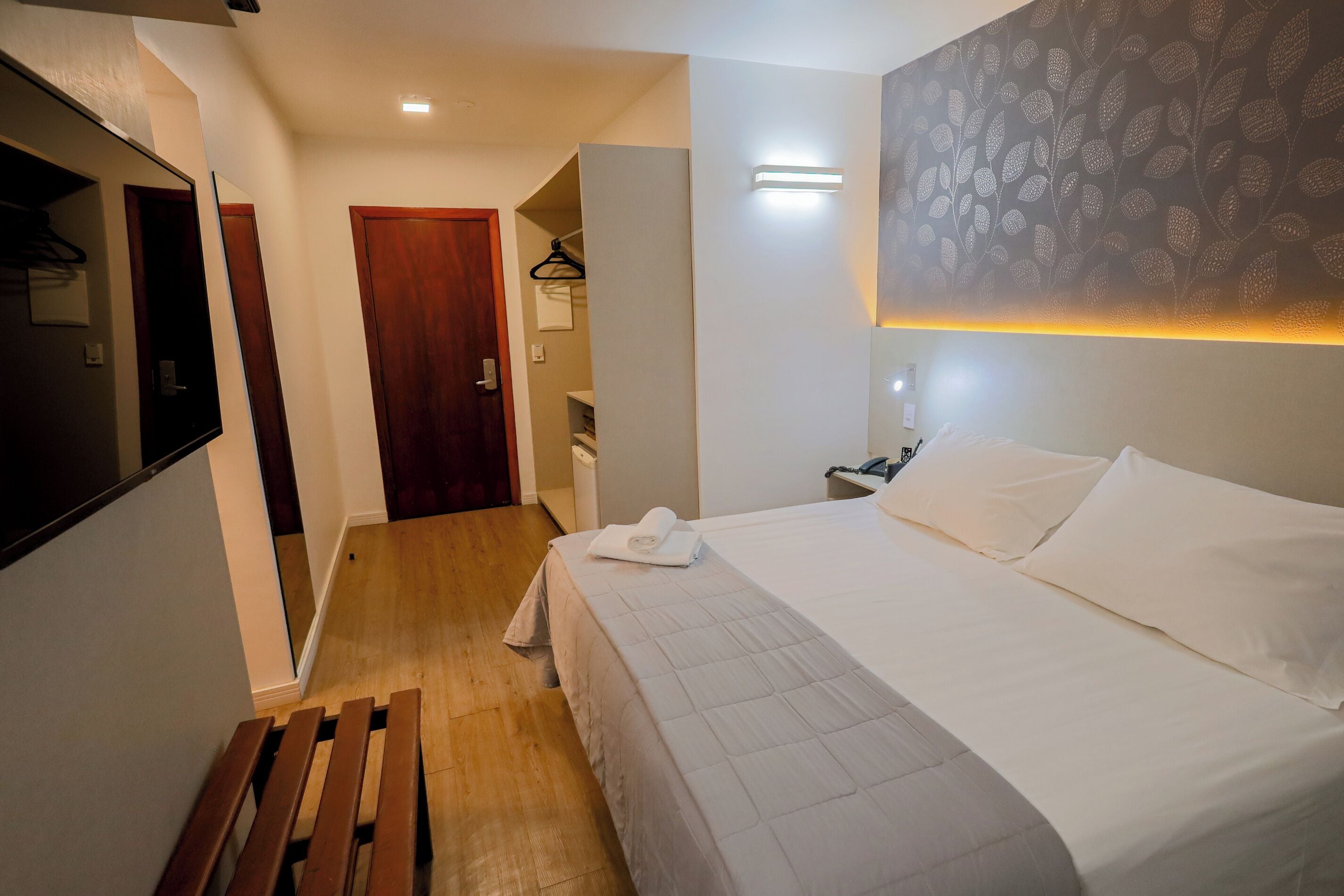 Quarto de hotel com cama, um banco de madeira, espelho, guarda-roupa e porta para um banheiro.