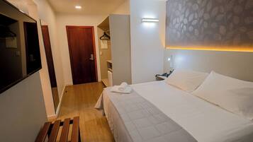 Quarto de hotel com cama, um banco de madeira, espelho, guarda-roupa e porta para um banheiro.