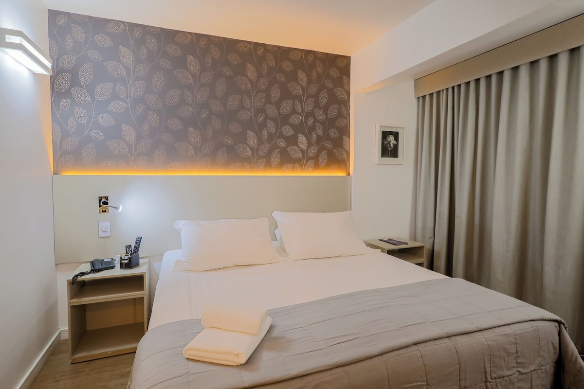 Quarto de hotel com cama, mesa de cabeceira, luminária, telefone e um quadro na parede.