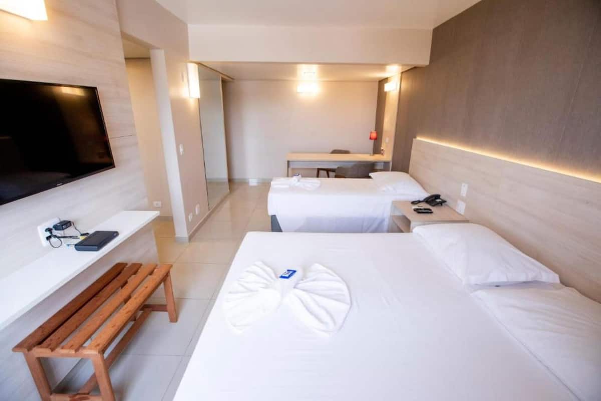 Quarto de hotel com televisão, um banco de madeira, duas camas com roupa branca e uma escrivaninha com abajur.