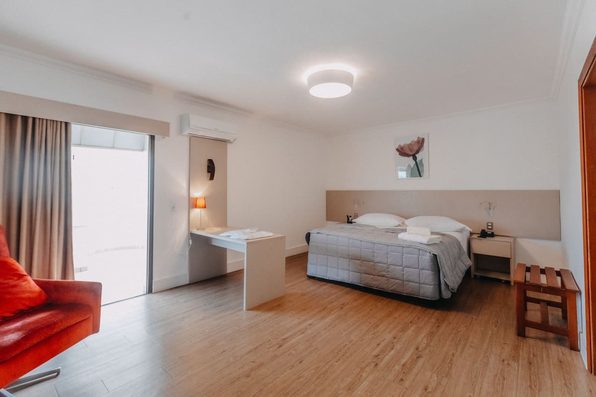 Quarto de hotel moderno com cama, mesas de cabeceira, escrivaninha, cadeira e vista através de uma porta de vidro.