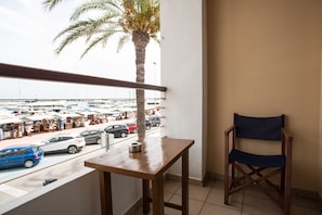 Uma varanda com mesa de madeira e cadeira azul, com vista para um estacionamento com carros e uma palmeira.