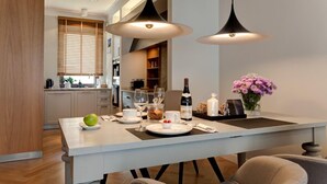 Uma área de jantar moderna com uma mesa branca posta para dois, incluindo uma garrafa de vinho, um vaso de flores e uma cesta de frutas.