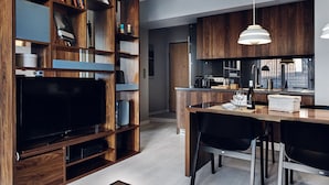 Uma sala de estar moderna com uma grande televisão de tela plana, estantes de madeira com livros e objetos decorativos, uma área de jantar com mesa e cadeiras de madeira, e uma cozinha com armários de madeira escura e iluminação pendente.