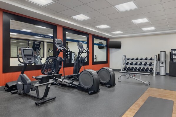 Uma academia com equipamentos de cardio, halteres e um espelho grande.