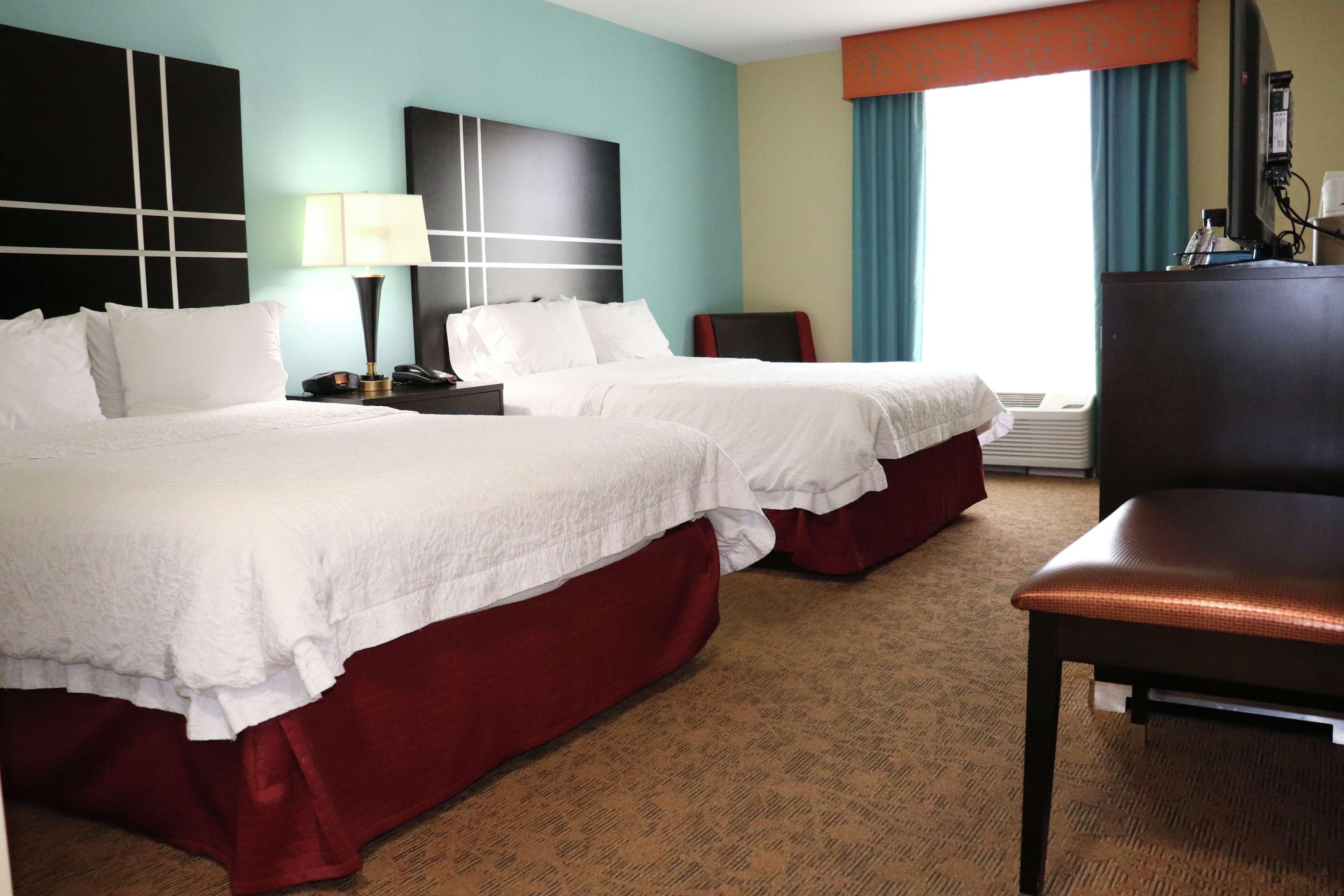 Quarto de hotel com duas camas, uma mesa, uma cadeira e uma janela grande com cortinas.