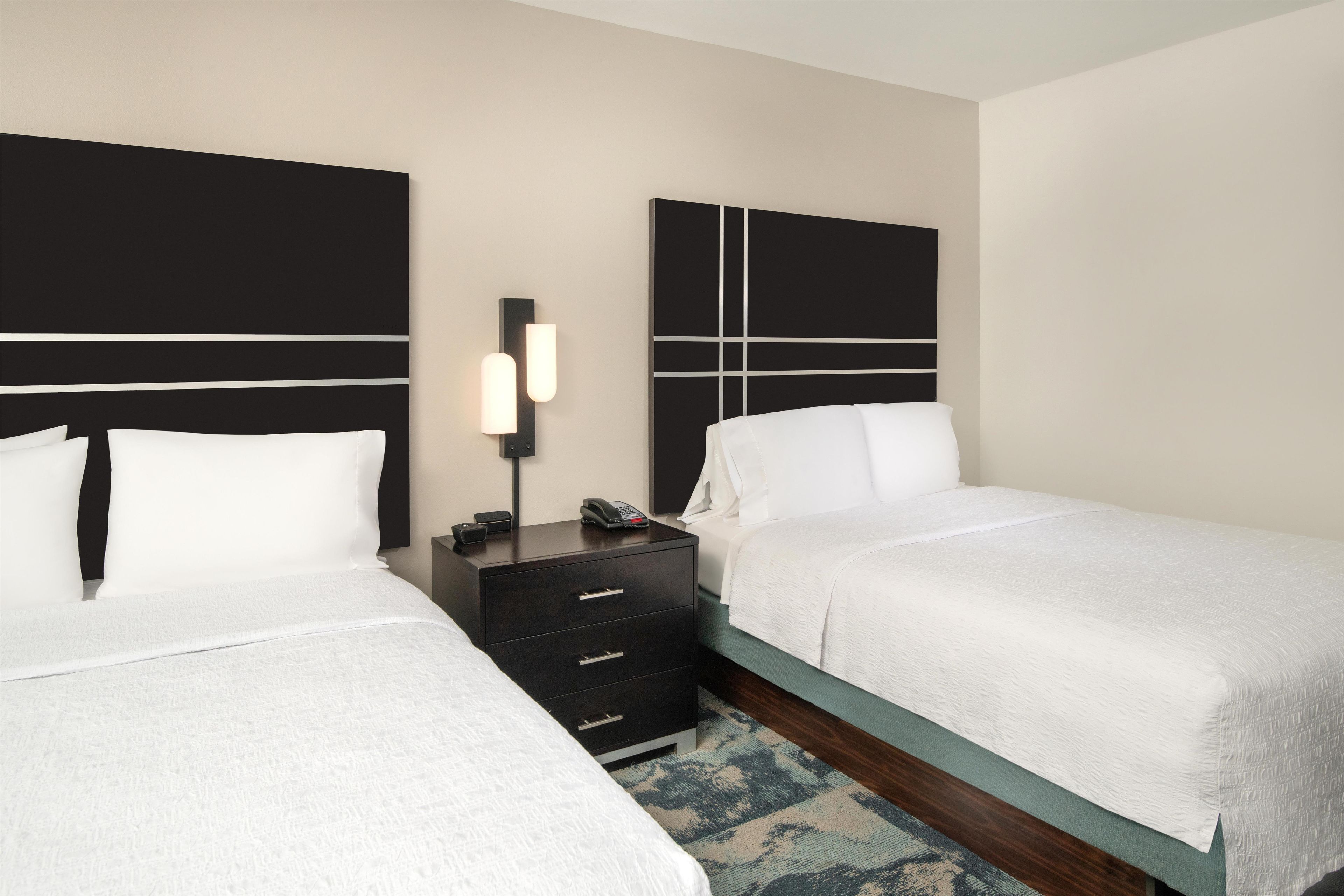 Quarto de hotel com duas camas, uma mesinha de cabeceira, um abajur e um telefone.