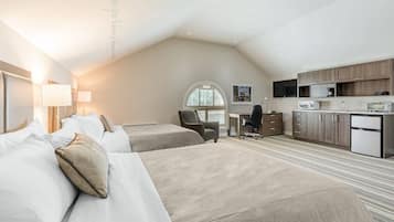 Quarto de hotel com duas camas, uma escrivaninha, uma cadeira, cozinha compacta e uma janela grande.