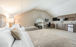 Quarto de hotel com duas camas, uma escrivaninha, uma cadeira, cozinha compacta e uma janela grande.