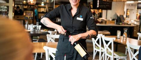 Uma pessoa servindo vinho em restaurante com cadeiras brancas e luminárias pendentes.
