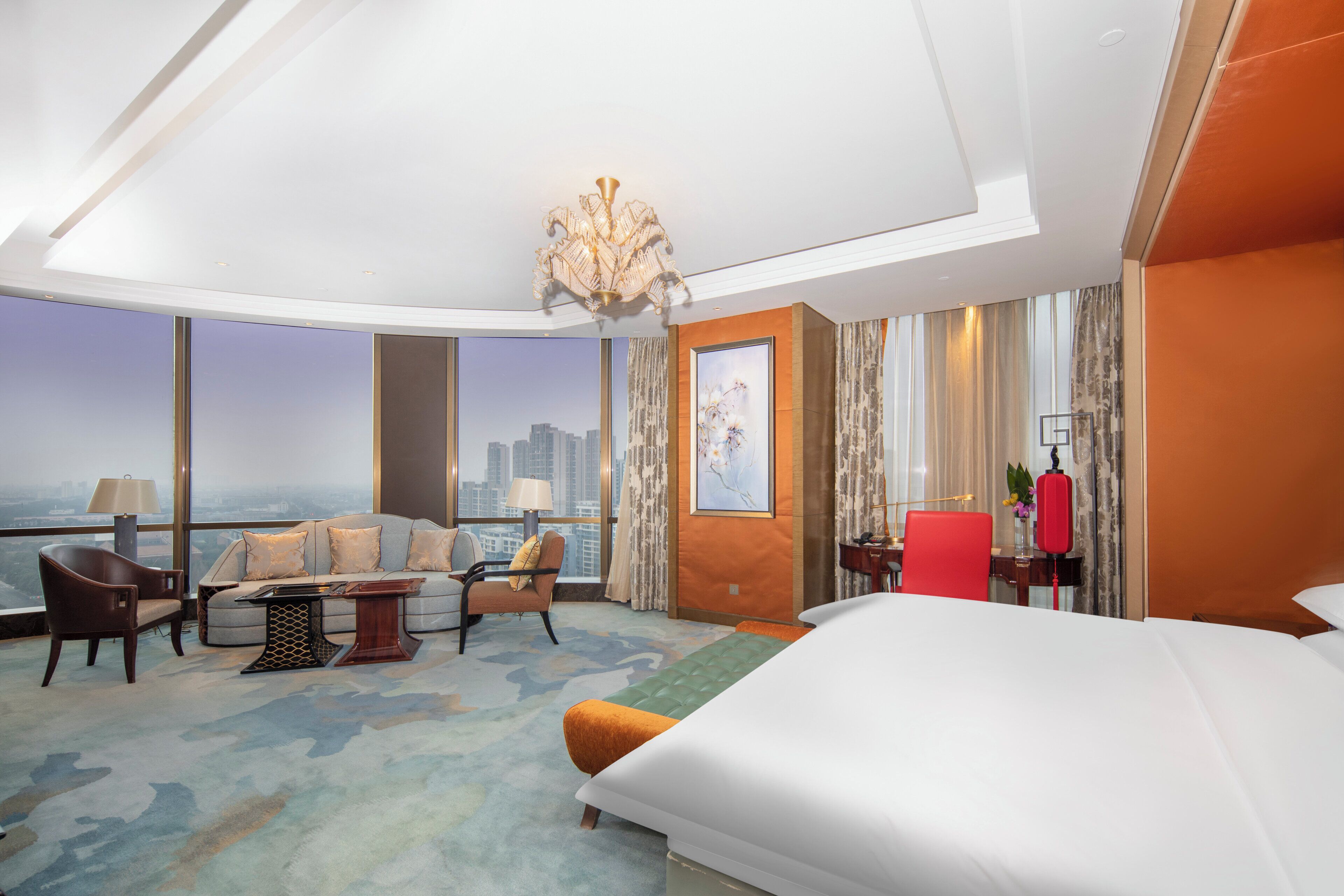 Quarto de hotel com uma cama grande, um sofá, uma cadeira, uma mesinha e vista para a cidade através de amplas janelas.