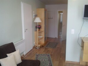 Um quarto com soalho de madeira, porta branca, sofá castanho, candeeiro de chão e um pequeno armário de madeira.