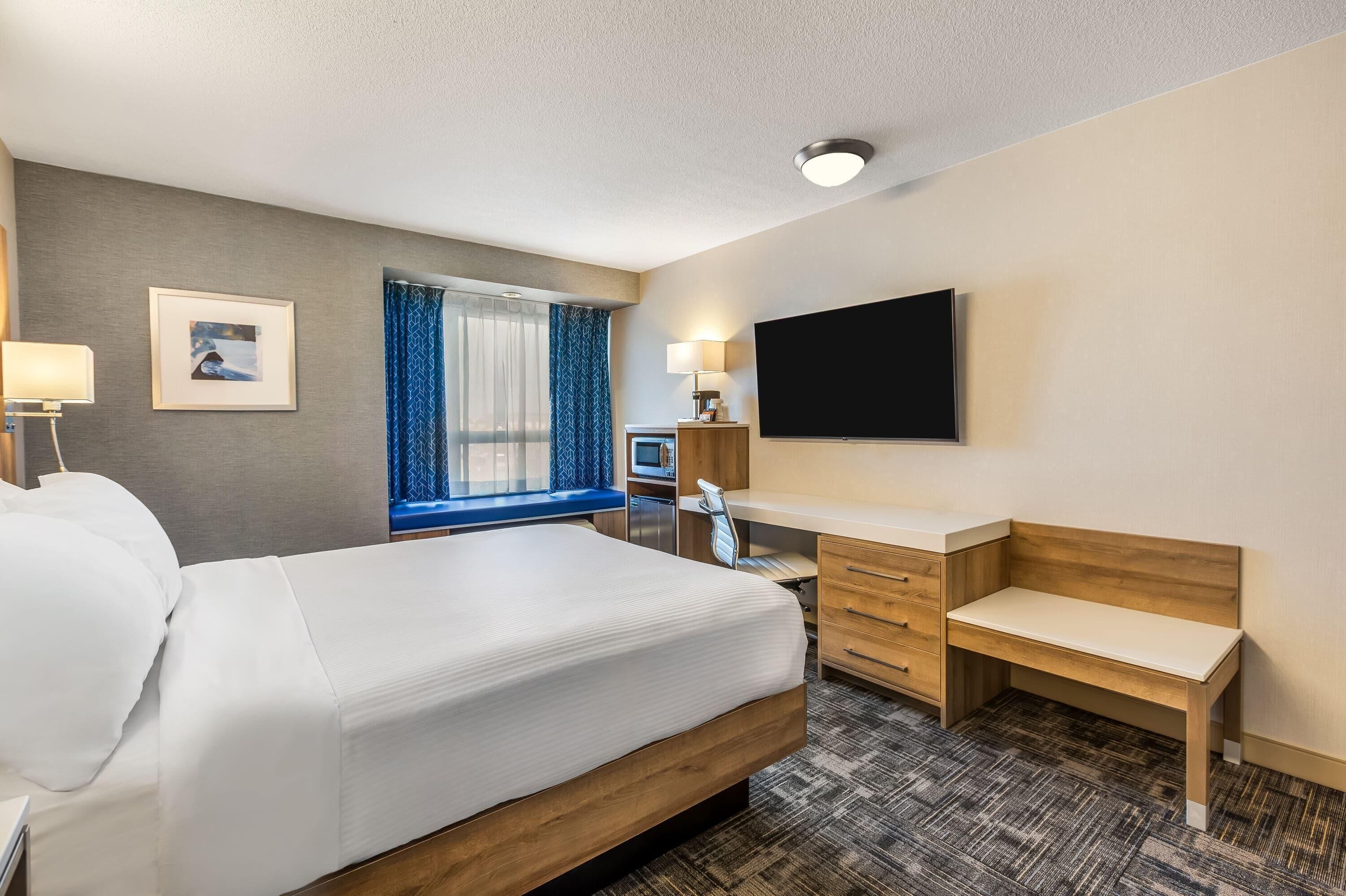 Quarto de hotel com cama, escrivaninha, cadeira, televisão e janela com cortinas.