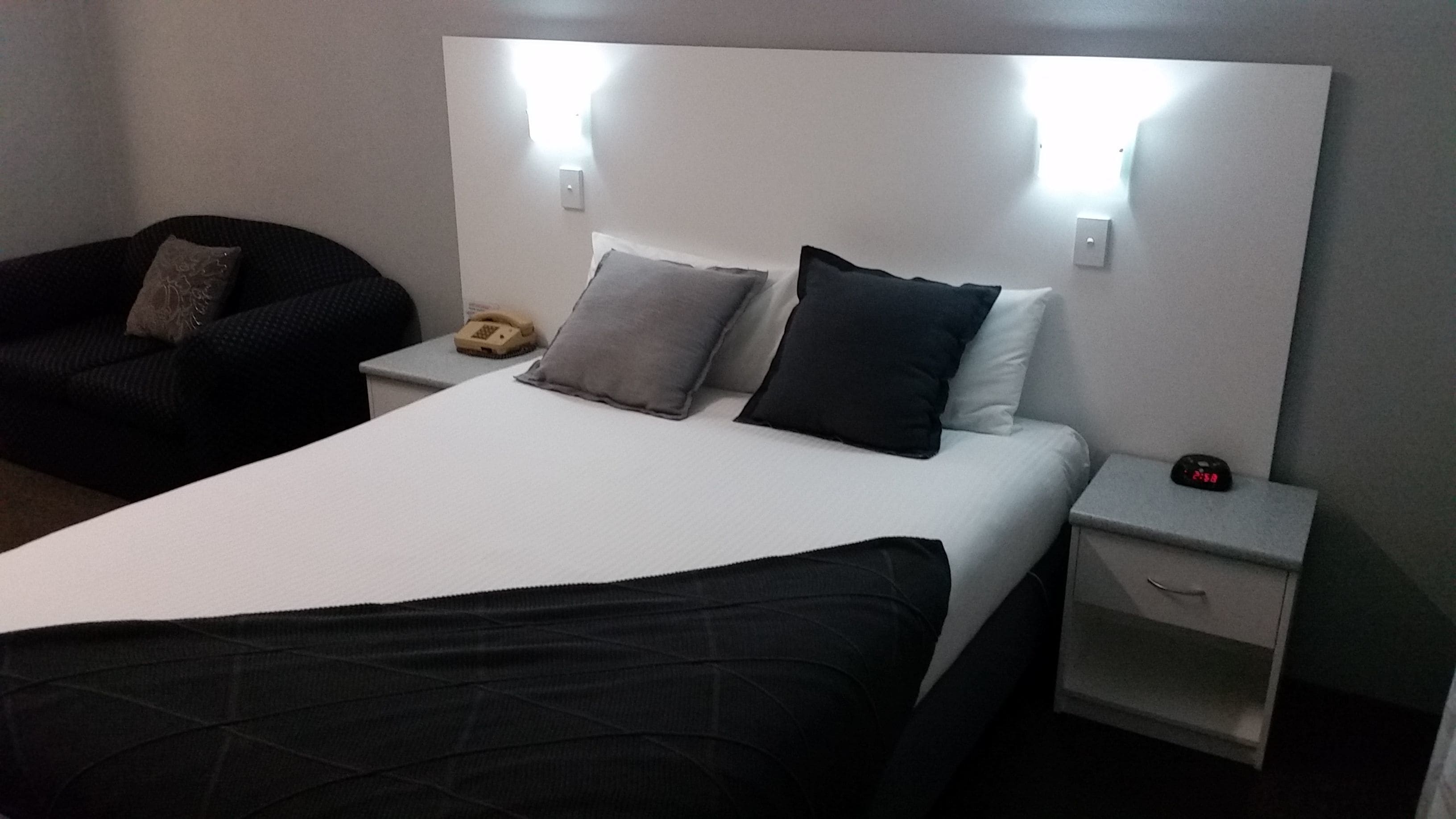 Quarto de hotel com cama grande, duas mesas de cabeceira, uma cadeira e uma mesinha de noite com relógio.