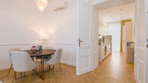 Uma sala de jantar com mesa, cadeiras e um candelabro. Uma cozinha é visível através de uma porta aberta.