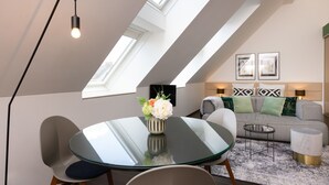 Uma sala de estar moderna com uma mesa de jantar de vidro, duas cadeiras, um sofá e uma mesa de centro. Há claraboias acima da mesa, e um abajur à esquerda.