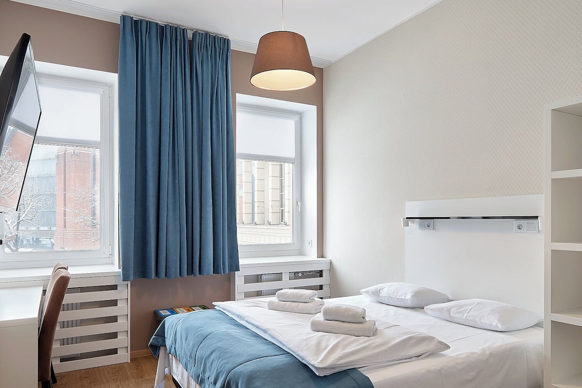 Quarto de hotel moderno com piso de madeira, cama com lençóis brancos, escrivaninha com cadeira e uma grande janela com cortinas azuis.