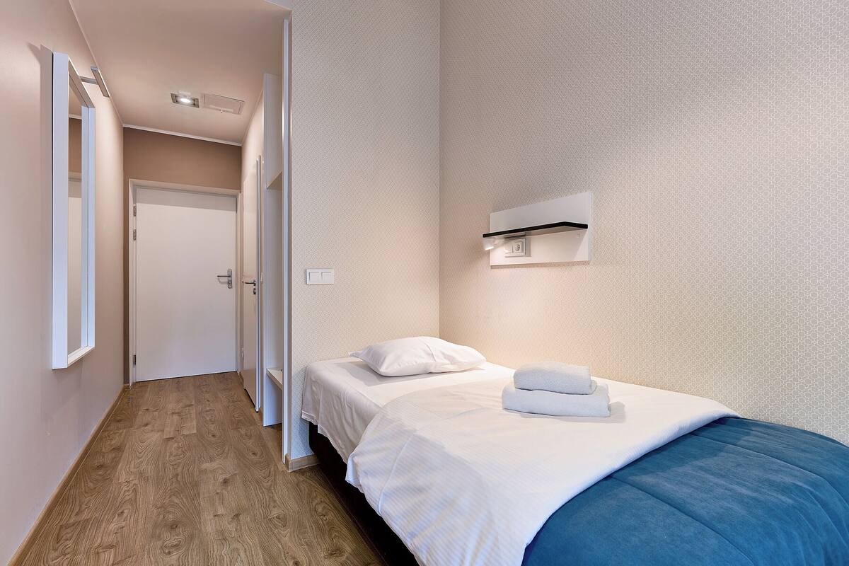 Quarto de hotel com uma cama, mesa de cabeceira, prateleira na parede e porta que dá para outro cômodo.