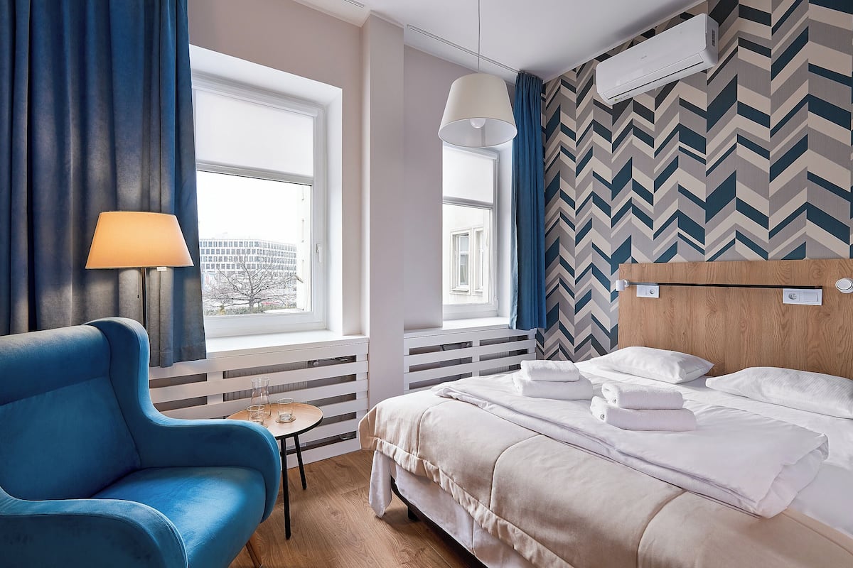 Quarto de hotel com cama, uma poltrona azul, uma mesinha e janela com cortinas.