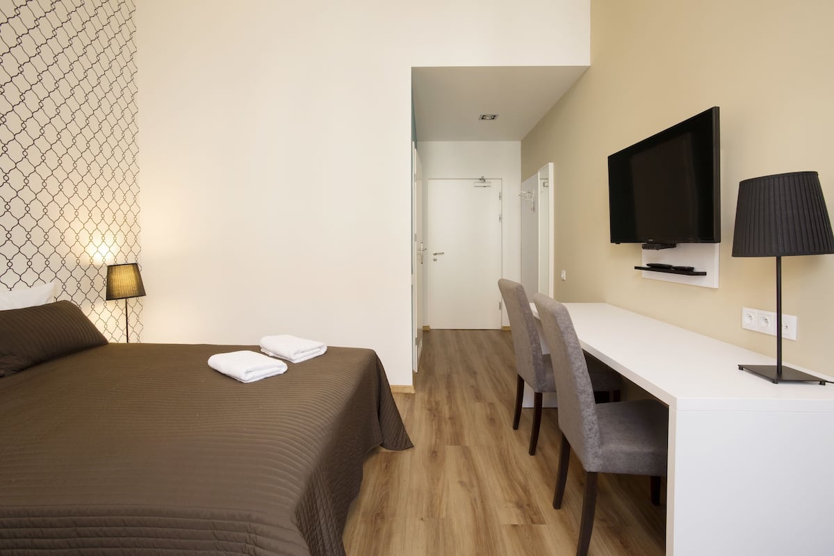 Quarto de hotel com cama, escrivaninha, cadeira, televisão e abajur.