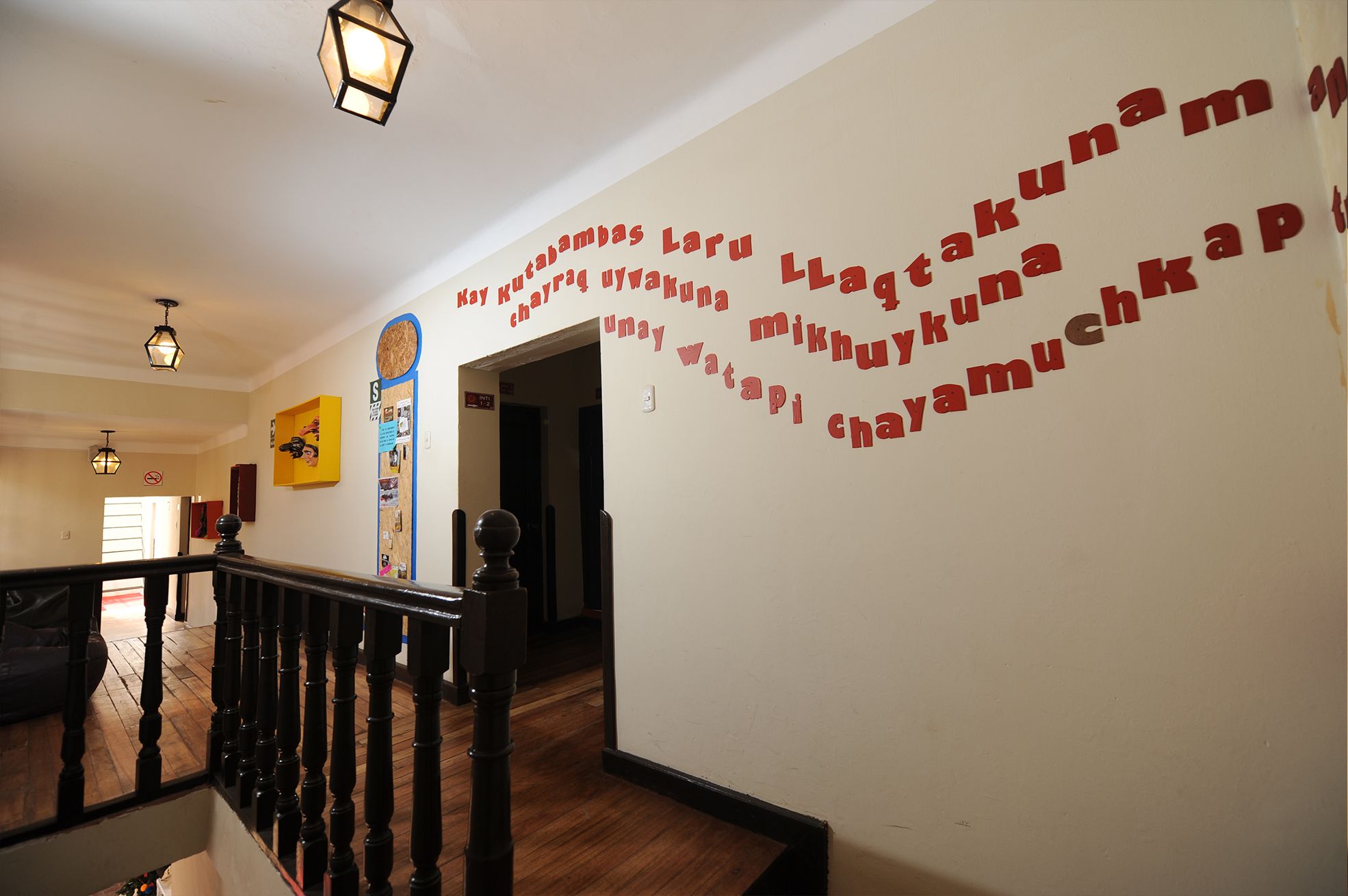 Um corredor com escadas de madeira, um mural com fotografias e uma parede com texto em língua estrangeira.