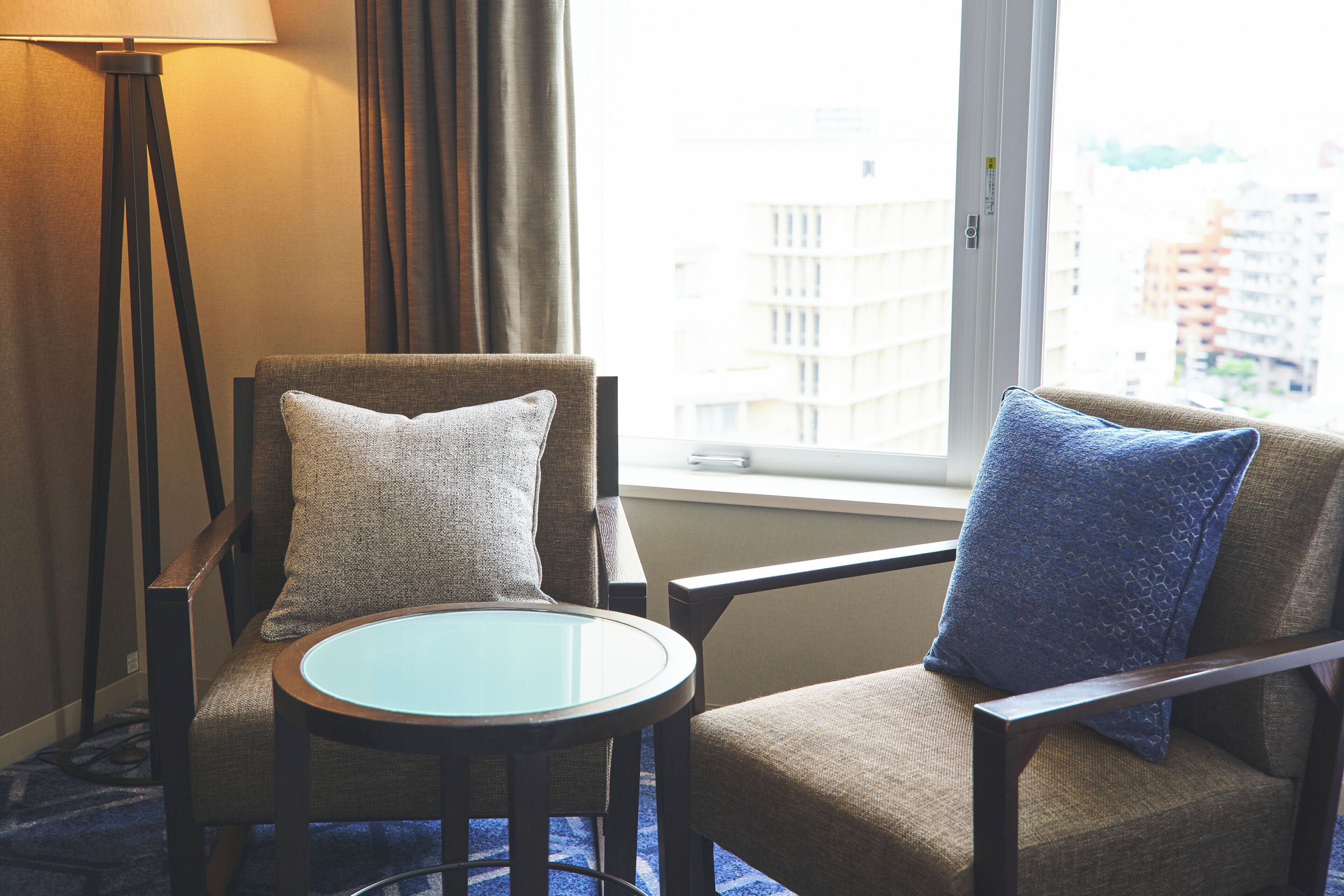 Quarto de hotel com duas poltronas, uma mesa pequena, abajur e janela com vista para a cidade.