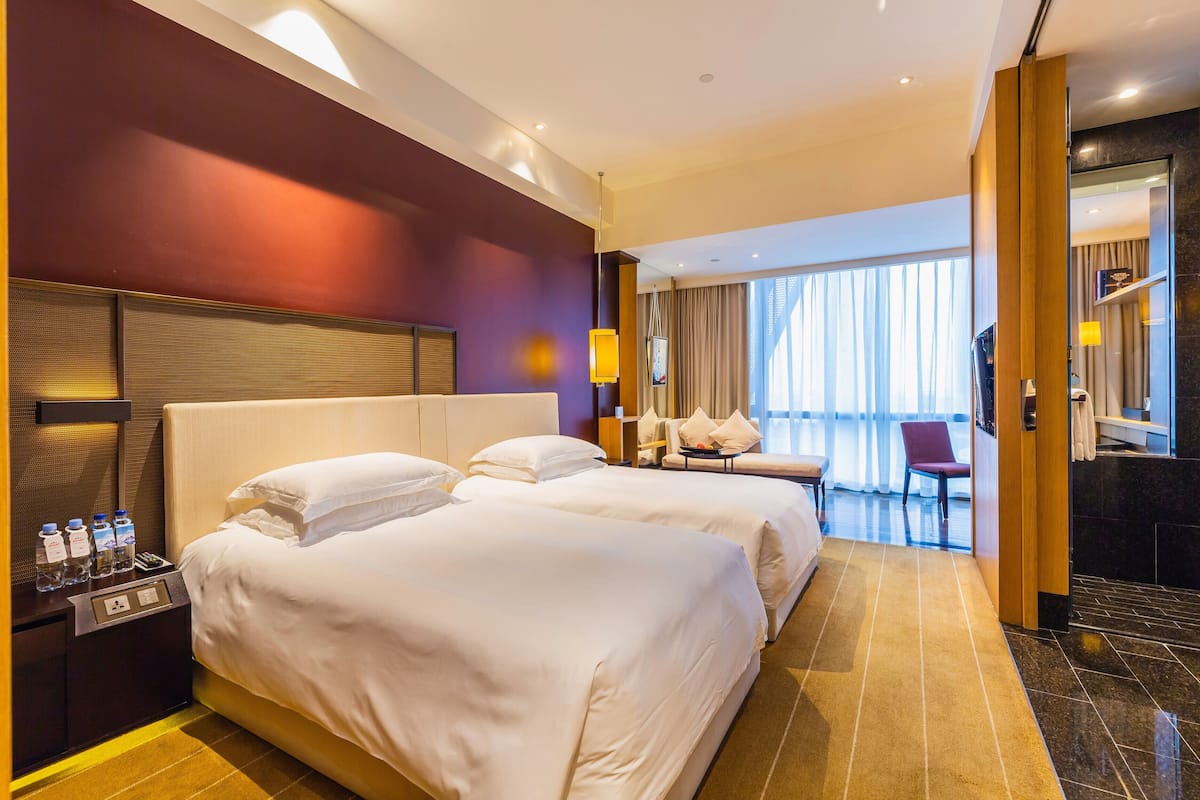 Quarto de hotel com duas camas, uma mesa de cabeceira com garrafas de água, uma cadeira e vista para o exterior através de grandes janelas.
