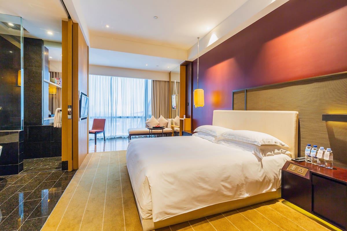 Quarto de hotel moderno com uma cama grande, uma escrivaninha com cadeira, televisão e vista para o exterior através de amplas janelas.