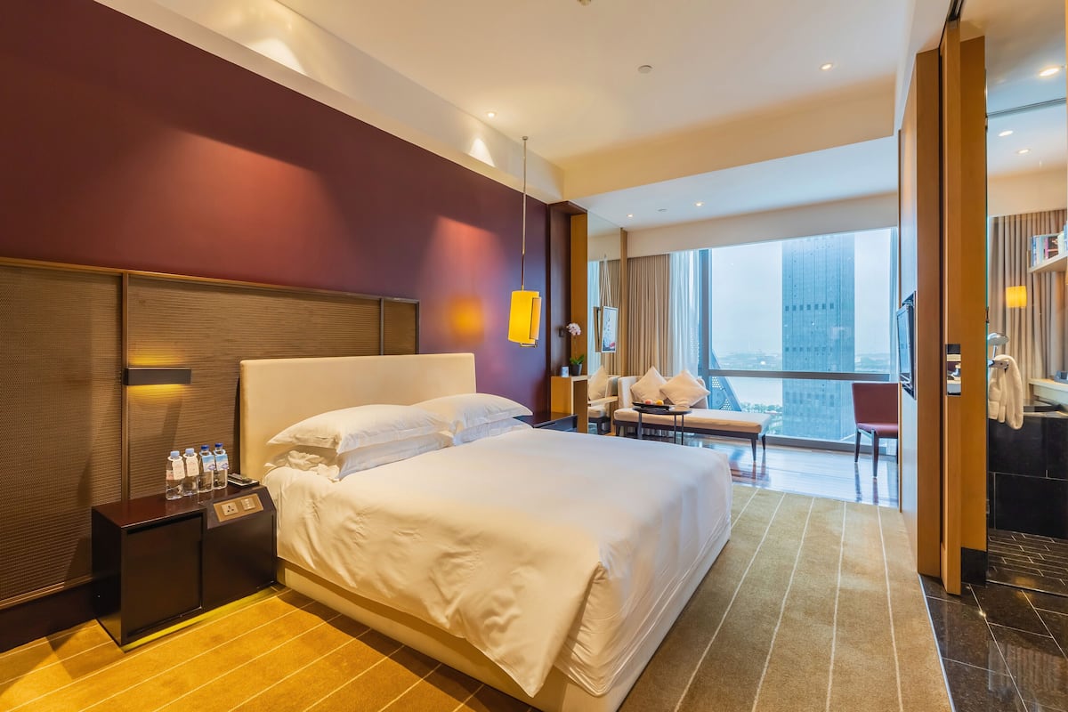 Quarto de hotel com uma cama grande, mesa de cabeceira com garrafas de água, uma cadeira, uma pequena mesa e vista para o horizonte urbano através de grandes janelas.