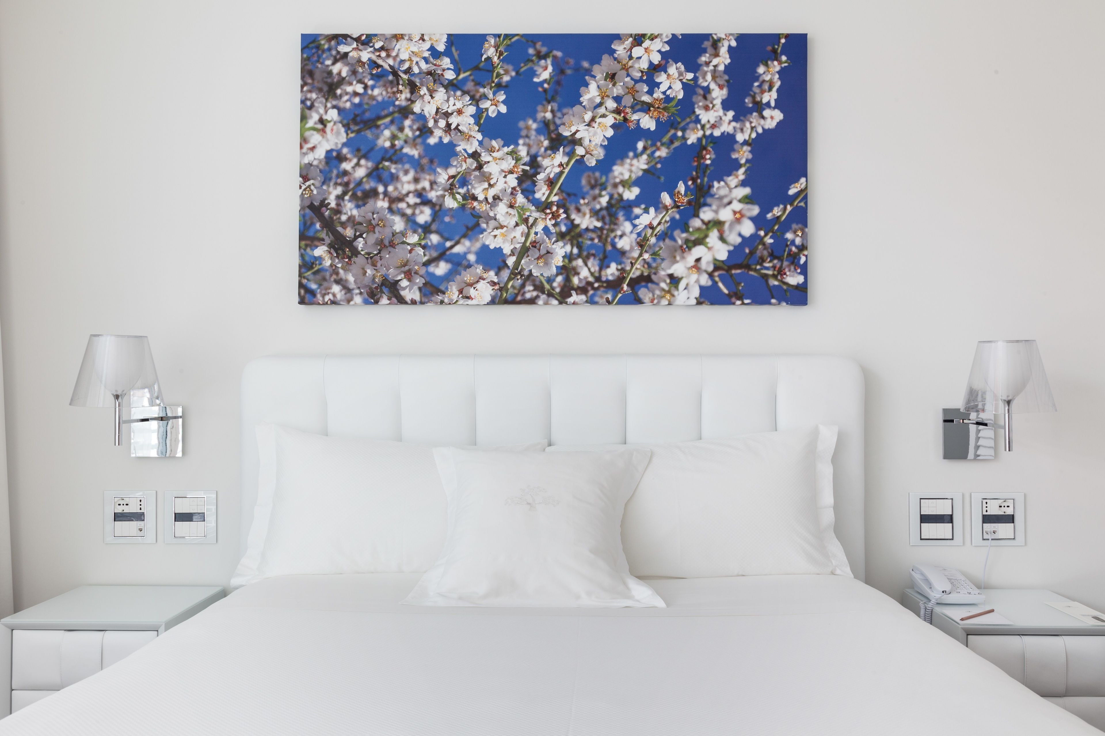 Quarto de hotel com uma cama grande, roupa de cama branca, um quadro de flores na parede, abajures e telefone ao lado da cama.