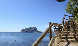 Uma paisagem costeira com uma escadaria de madeira que leva a uma plataforma rochosa, com vista para águas azuis calmas e uma montanha ao longe.
