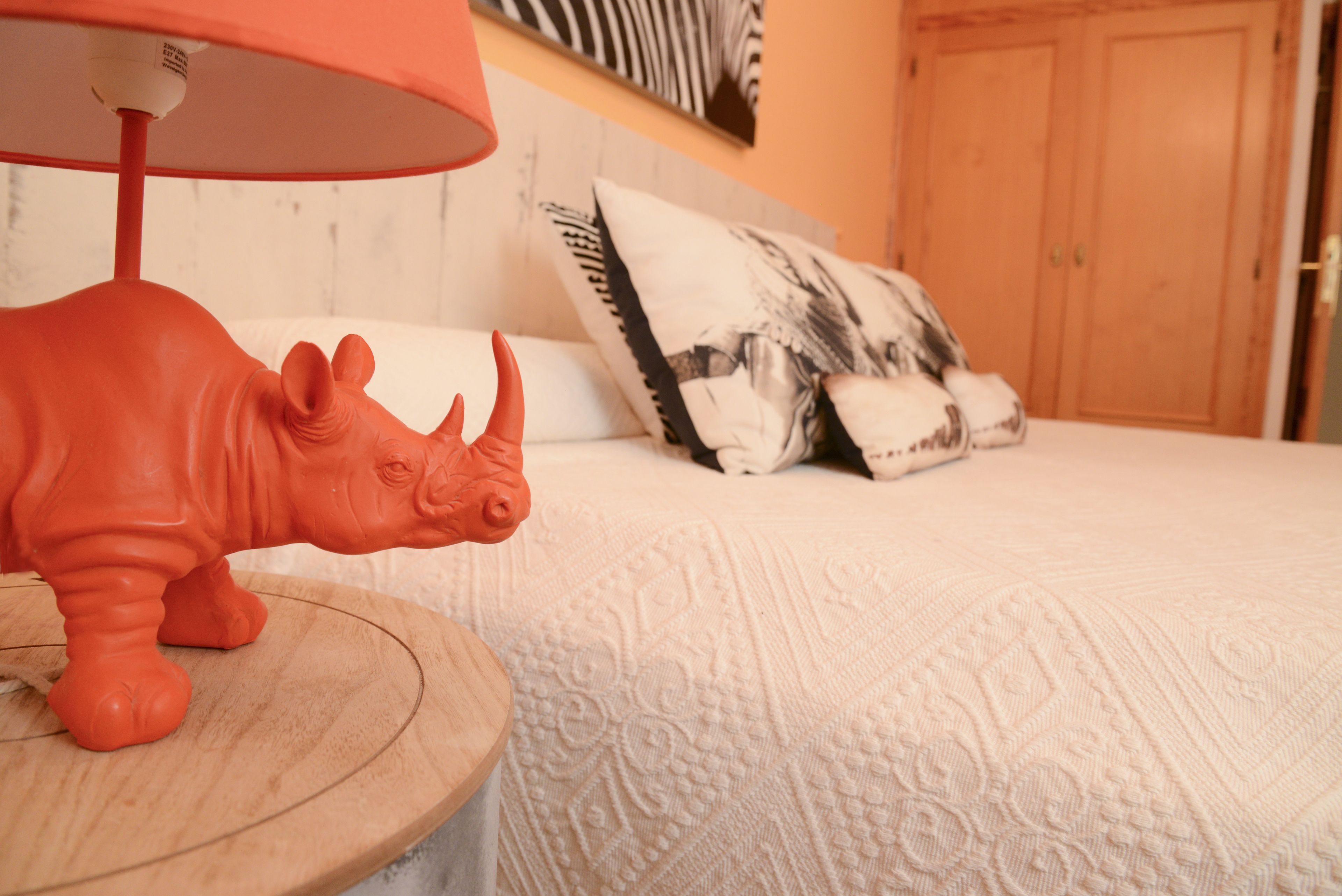 Uma cama com colcha branca texturizada, uma mesa de cabeceira redonda de madeira e uma estatueta de rinoceronte.