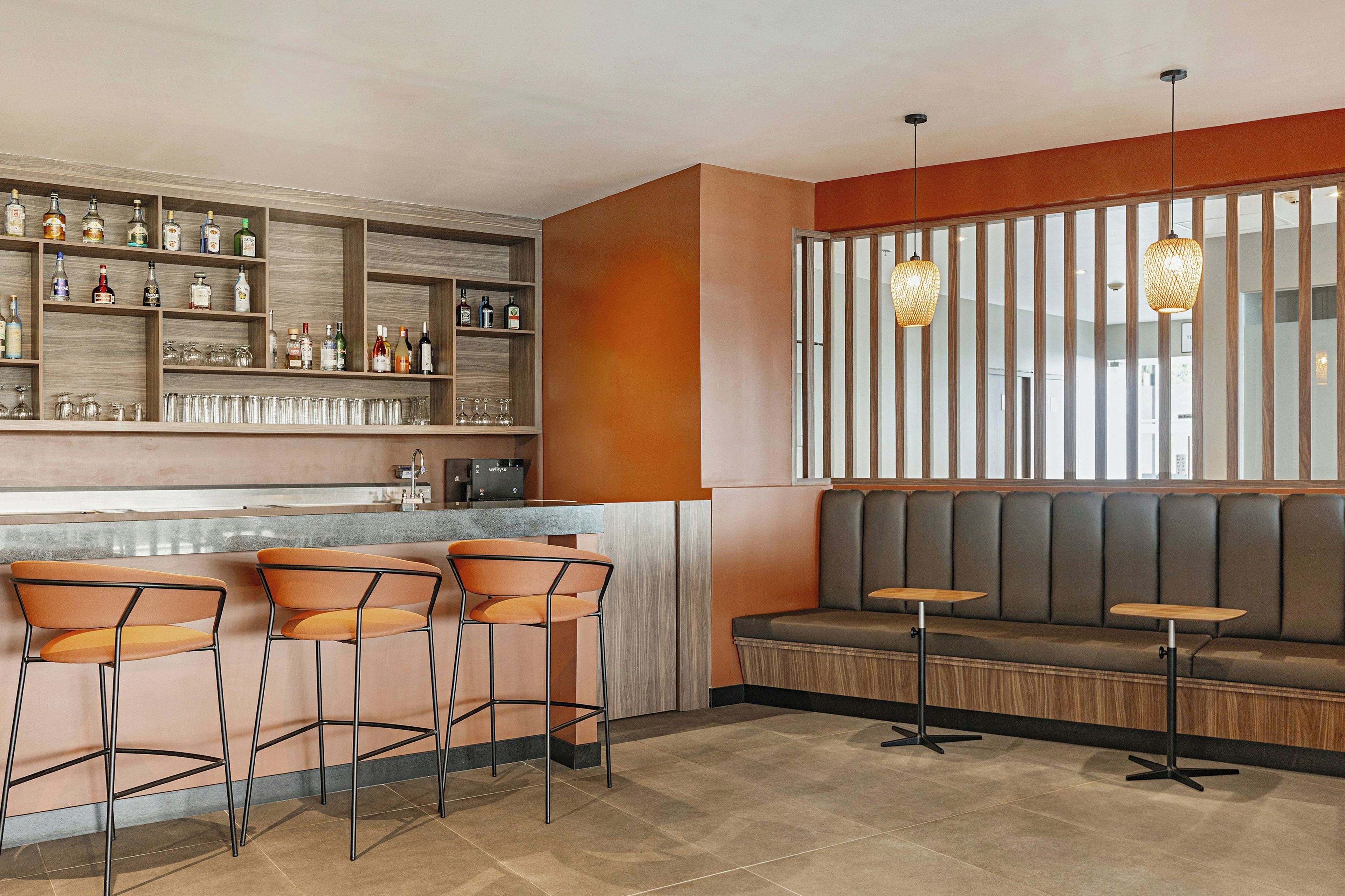 Uma área de bar com banquetas, balcão e prateleiras com garrafas e copos.
