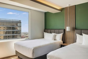 Quarto de hotel com duas camas, uma janela grande, vista para um edifício e uma parede de destaque verde.